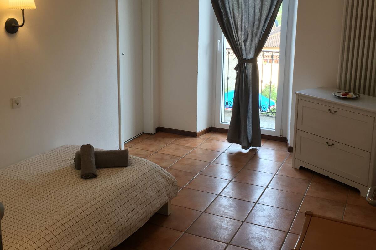 Quarto com cama, mesa de cabeceira, cômoda, janela com cortinas e luminária na parede.