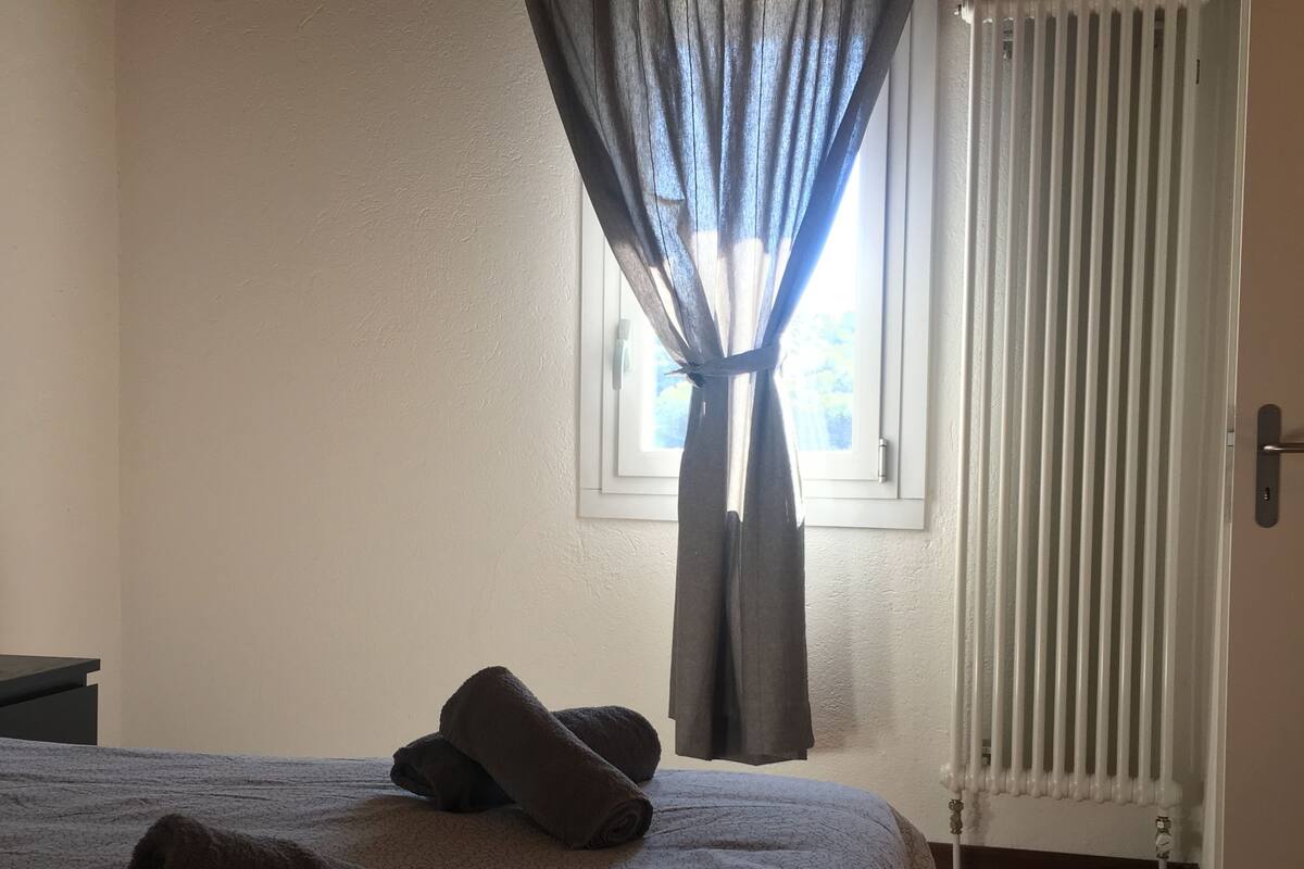Quarto com cama, toalhas dobradas, janela com cortinas, radiador e teto de madeira.