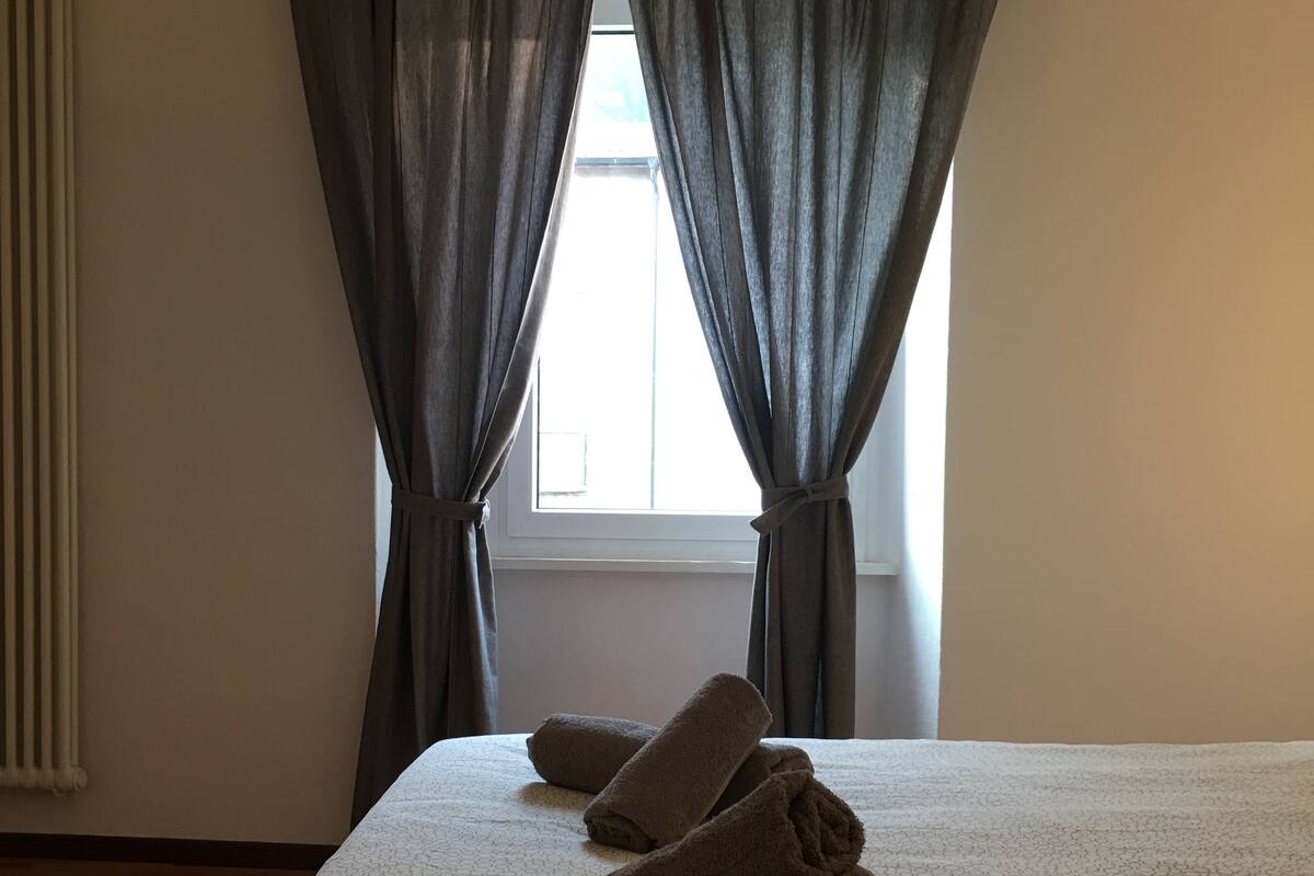 Quarto de hotel com cama, duas toalhas enroladas, radiador e janela com cortinas.
