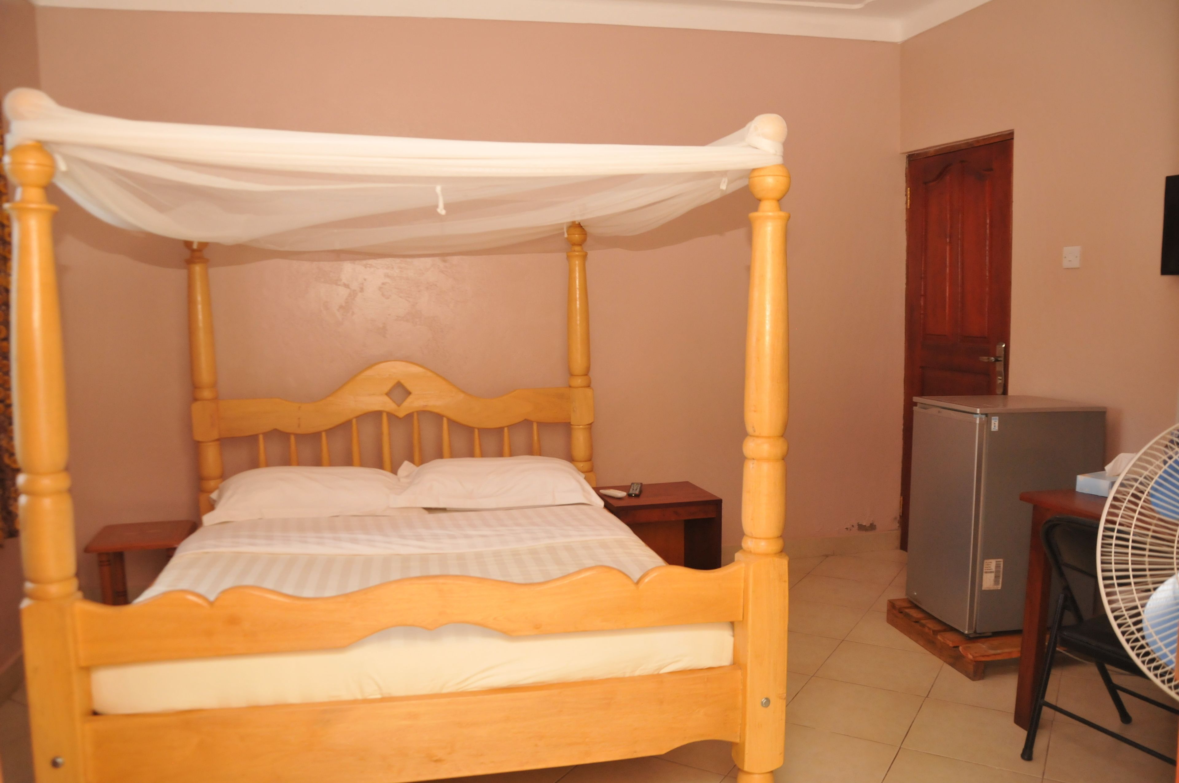 Uma cama com dossel e cortina, uma mesinha de cabeceira, um frigorífico e um ventilador.