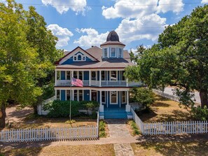 Uma casa de dois andares com varanda em volta, um mastro com a bandeira americana e uma cerca branca de piquete.