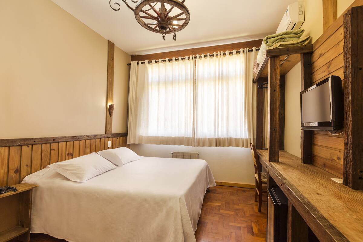 Quarto com cama e escrivaninha de madeira, televisão e janela com cortinas.