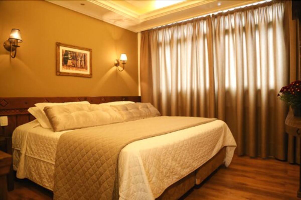 Quarto de hotel com uma cama grande, duas luzes de parede, um quadro emoldurado e uma janela com cortinas.