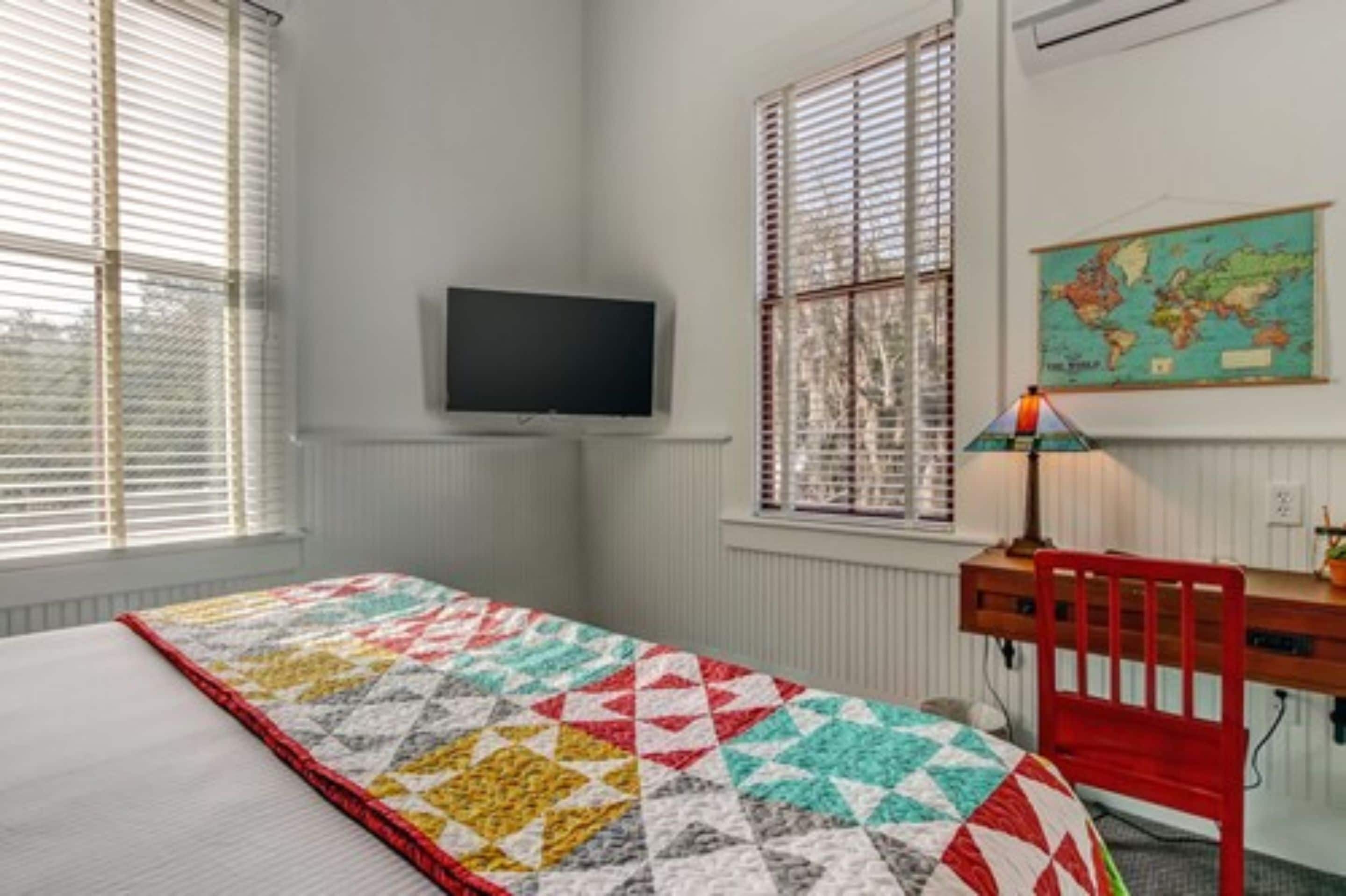Um quarto com uma cama, uma escrivaninha com cadeira, uma televisão e um mapa-múndi na parede.