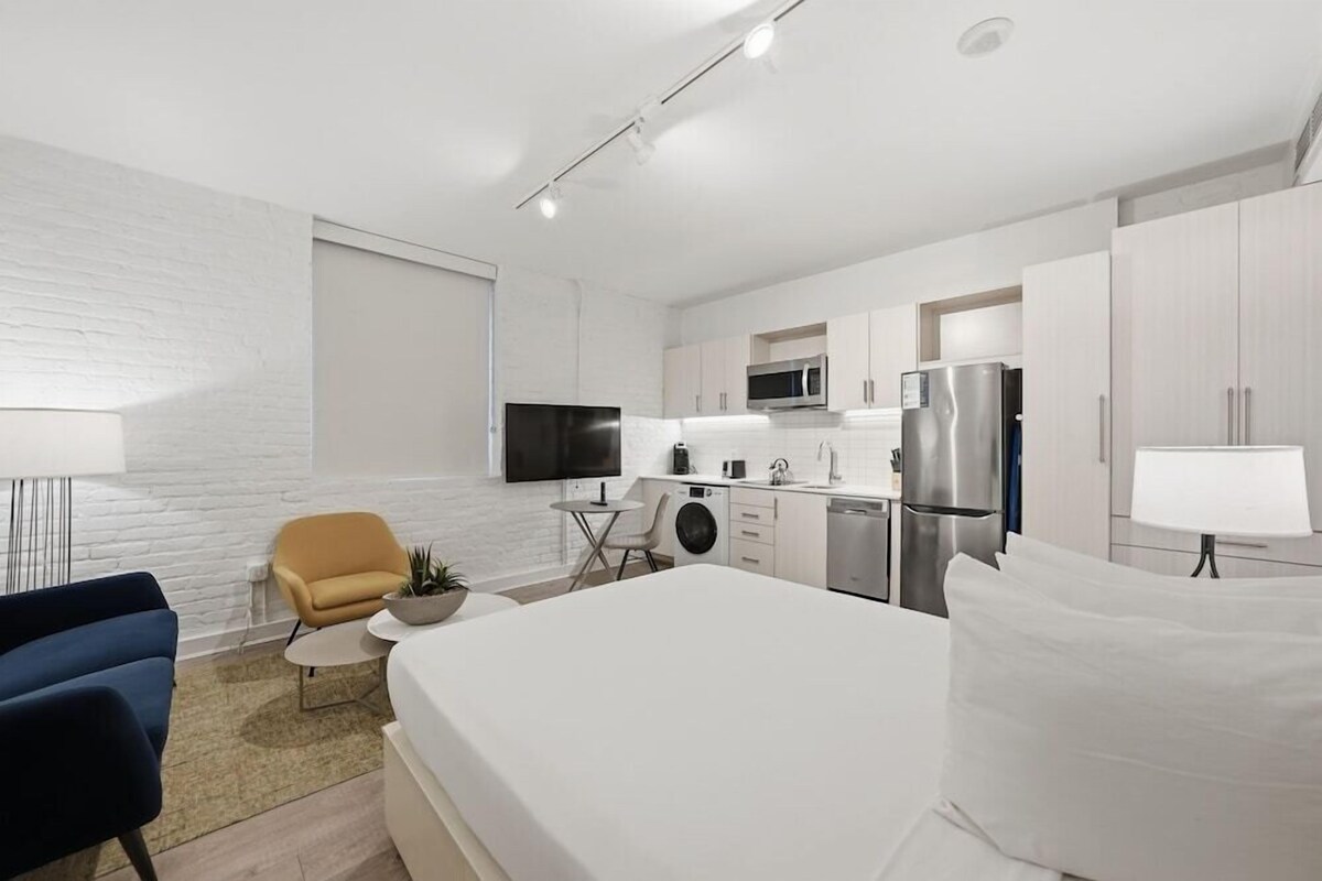 Quarto de hotel moderno com cama, sofá, pequena mesa de jantar, TV, área de cozinha equipada com micro-ondas, frigorífico e máquina de lavar roupa.