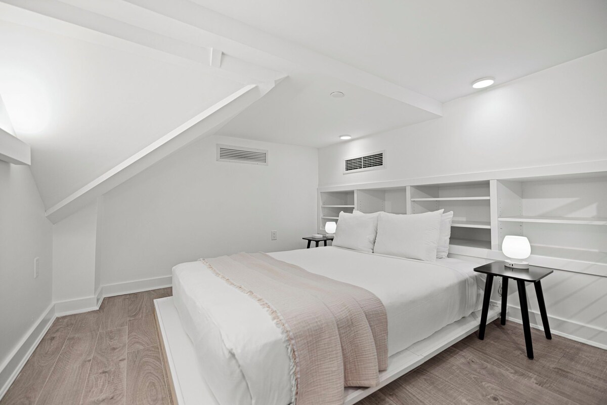 Quarto moderno com teto inclinado, cama com roupa de cama branca, mesa de cabeceira com abajur e nichos embutidos.