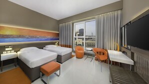 Quarto de hotel com duas camas, uma escrivaninha, uma cadeira e uma janela grande com vista para a cidade.