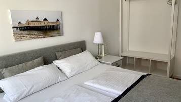 Cama bem arrumada com lençóis brancos e cabeceira cinzenta, mesa de cabeceira com abajur e quadro na parede retratando um edifício.
