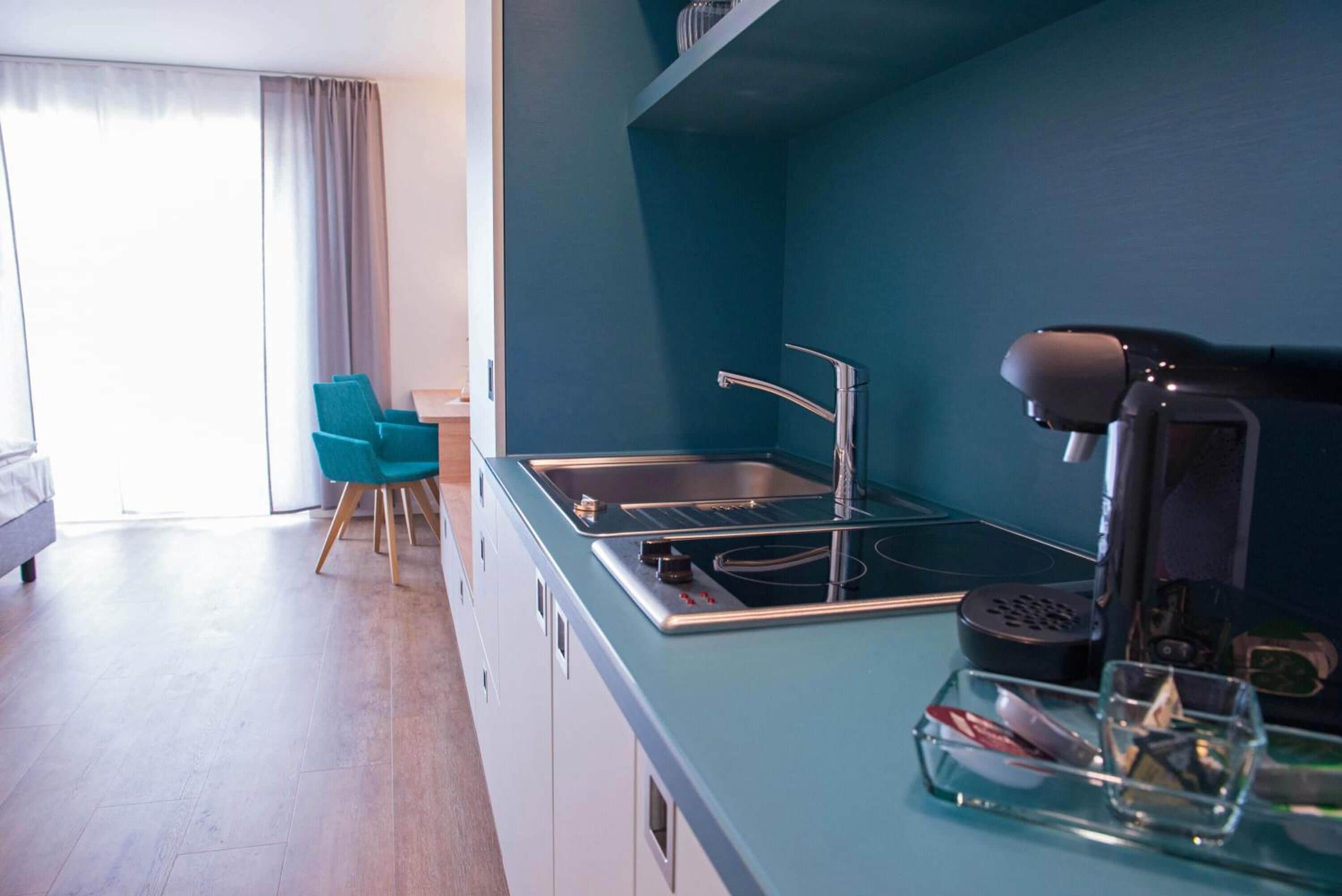 Uma kitchenette compacta com pia, fogão e cafeteira, bancada cor verde-água e uma cadeira próxima a uma janela com cortinas.