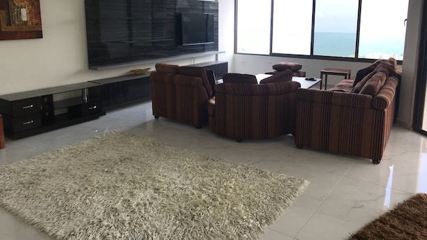 Uma sala de estar moderna com uma televisão de tela plana grande, um sofá seccional e uma janela ampla com vista para o oceano.
