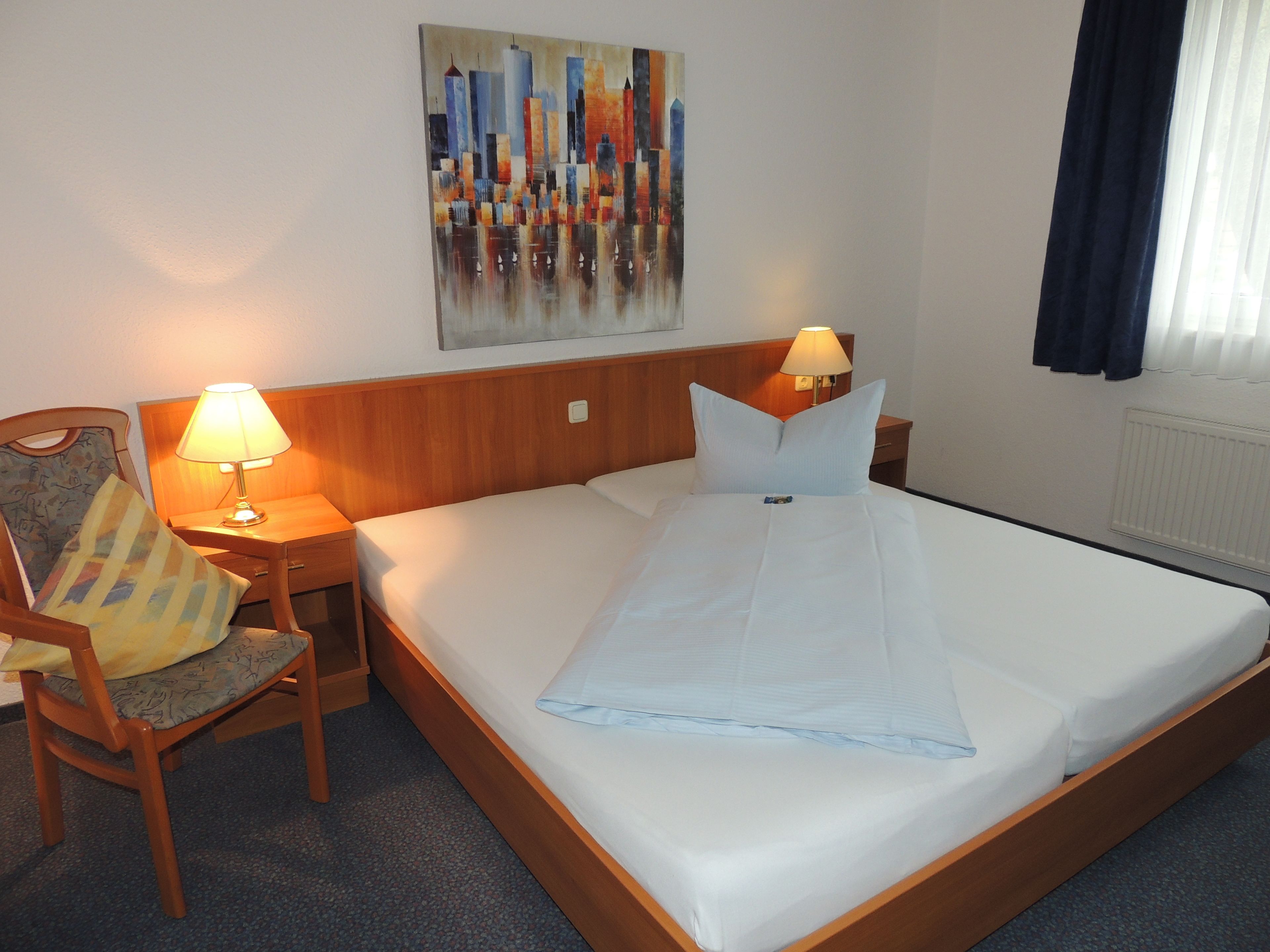 Quarto de hotel com uma cama de solteiro, duas luminárias de cabeceira, uma poltrona com almofada, um quadro na parede e uma janela com cortinas.