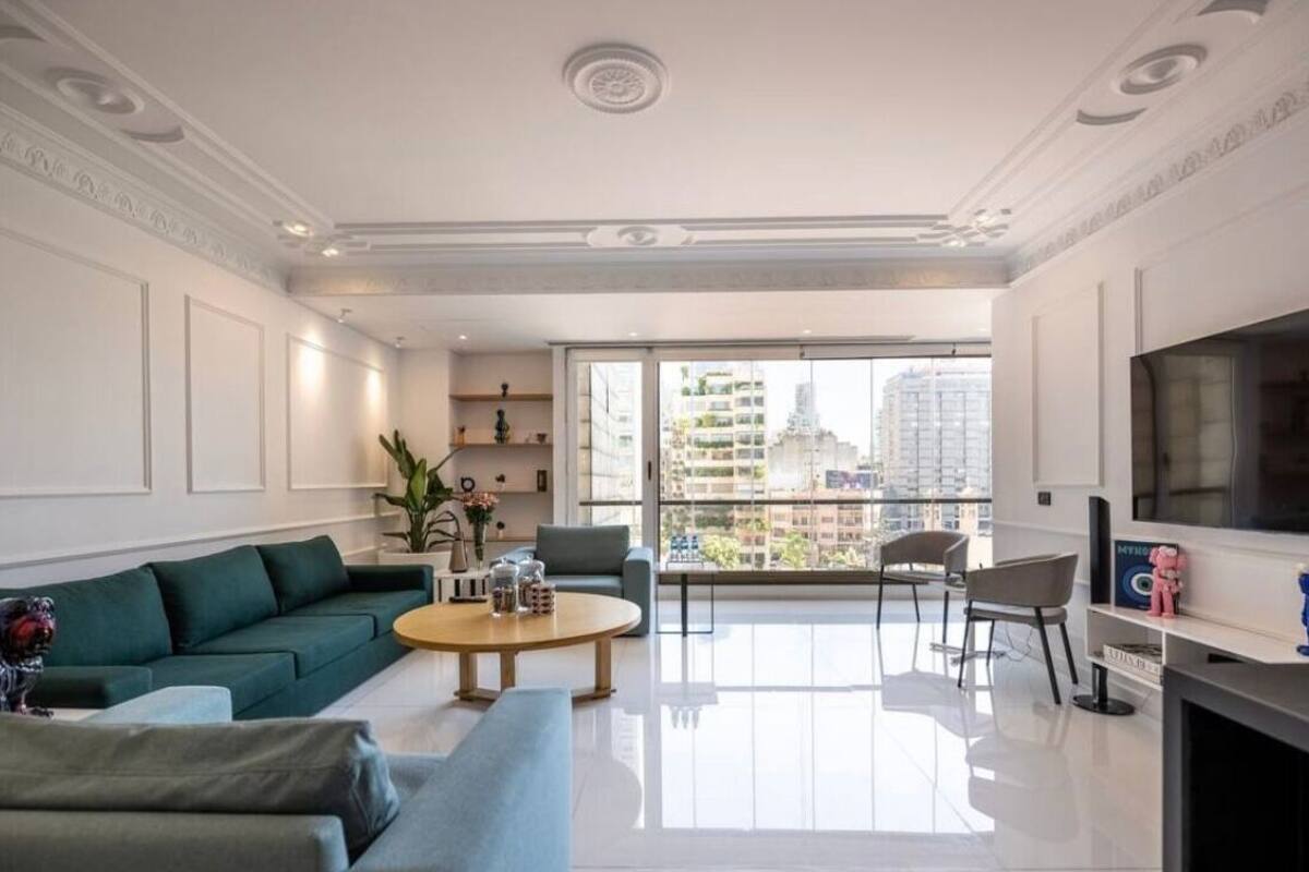 Sala de estar moderna com uma televisão grande, sofás verdes, uma mesa de centro e vista para a cidade através de amplas janelas.