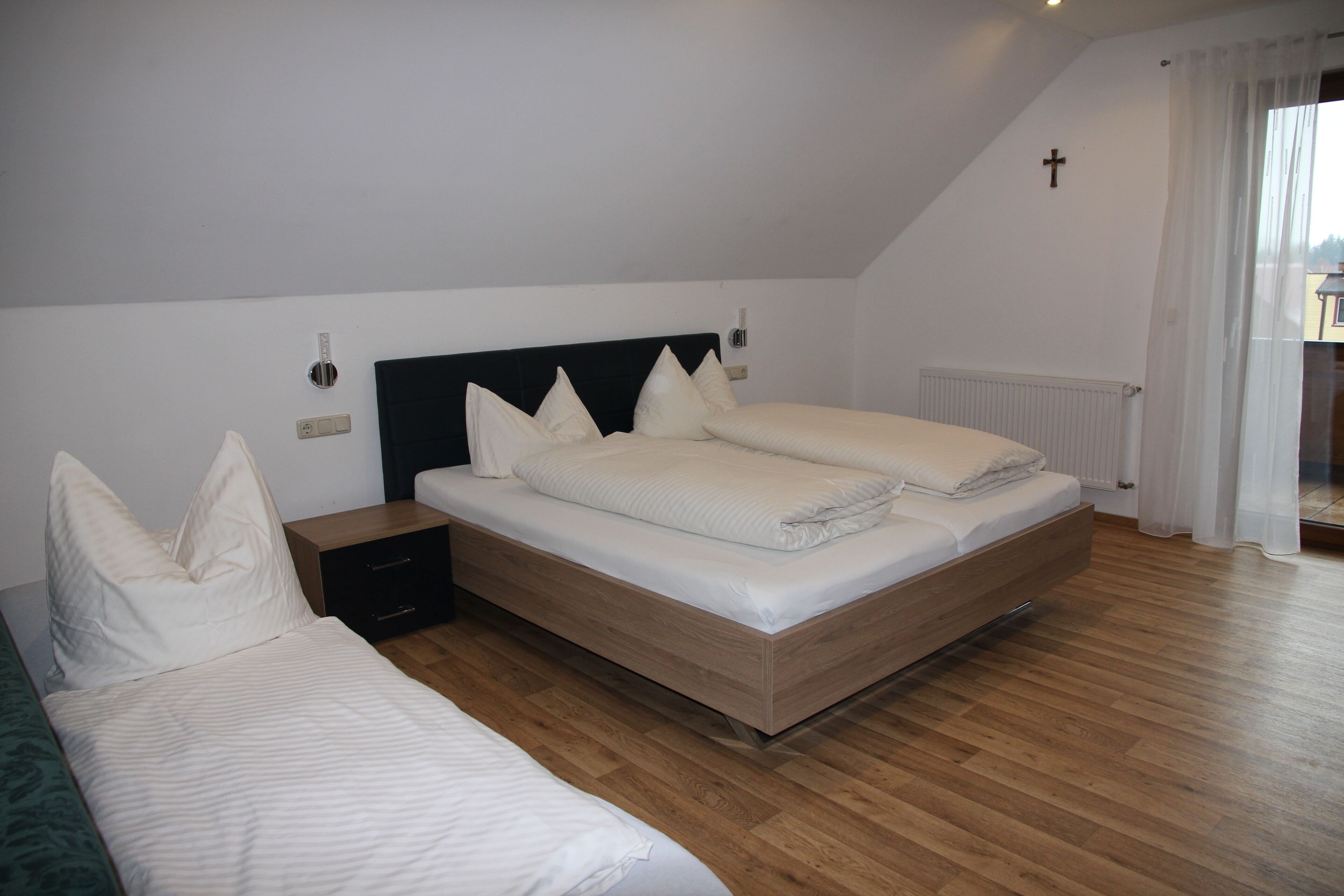 Quarto com uma cama grande, uma cama menor, piso de madeira, mesa de cabeceira, luz acoplada à parede e uma janela com cortinas.