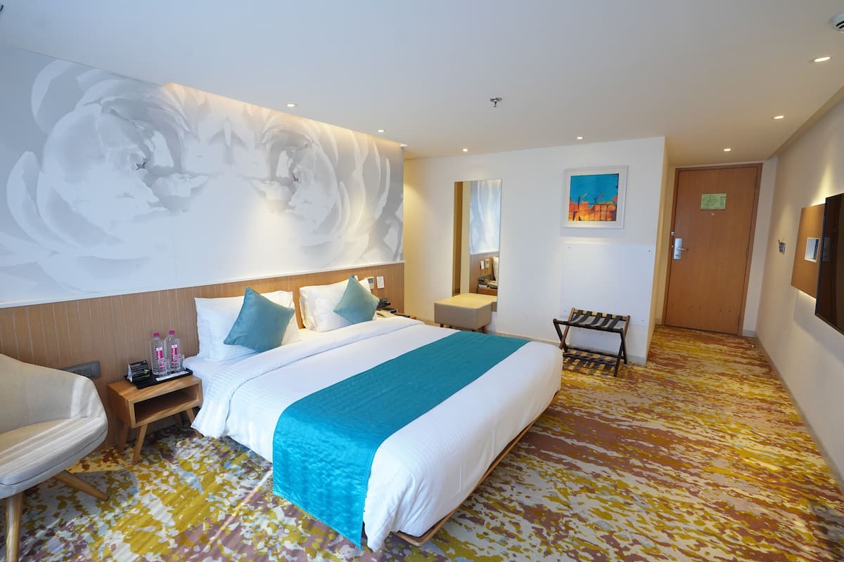 Quarto de hotel com uma cama grande, uma mesa de cabeceira com abajur, uma cadeira, um banco e um quadro abstrato grande na parede.