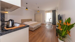 Quarto de hotel moderno com cozinha compacta, uma cama, área de refeições e uma janela grande com cortinas.