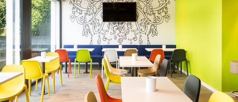 Uma área de jantar bem iluminada, com cadeiras e mesas coloridas, um grande mural na parede e uma TV de tela plana fixada na parede.