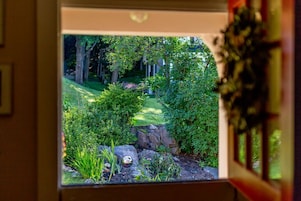 Vista pela janela de um jardim com pedras, arbustos e árvores.