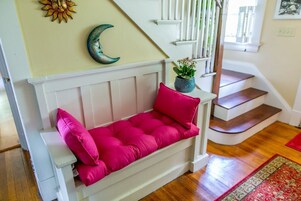 Um banco branco com almofadas rosa vibrantes, uma pequena planta em vaso e arte decorativa na parede, incluindo um sol e uma lua crescente.