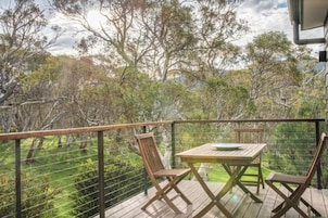 Um deck de madeira com mesa e cadeiras, cercado por guarda-corpo de metal, com vista para uma paisagem verde e exuberante com árvores.