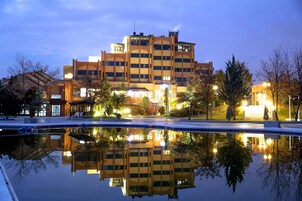 Um hotel com vários andares refletido em águas calmas, cercado por árvores e uma rua bem iluminada.