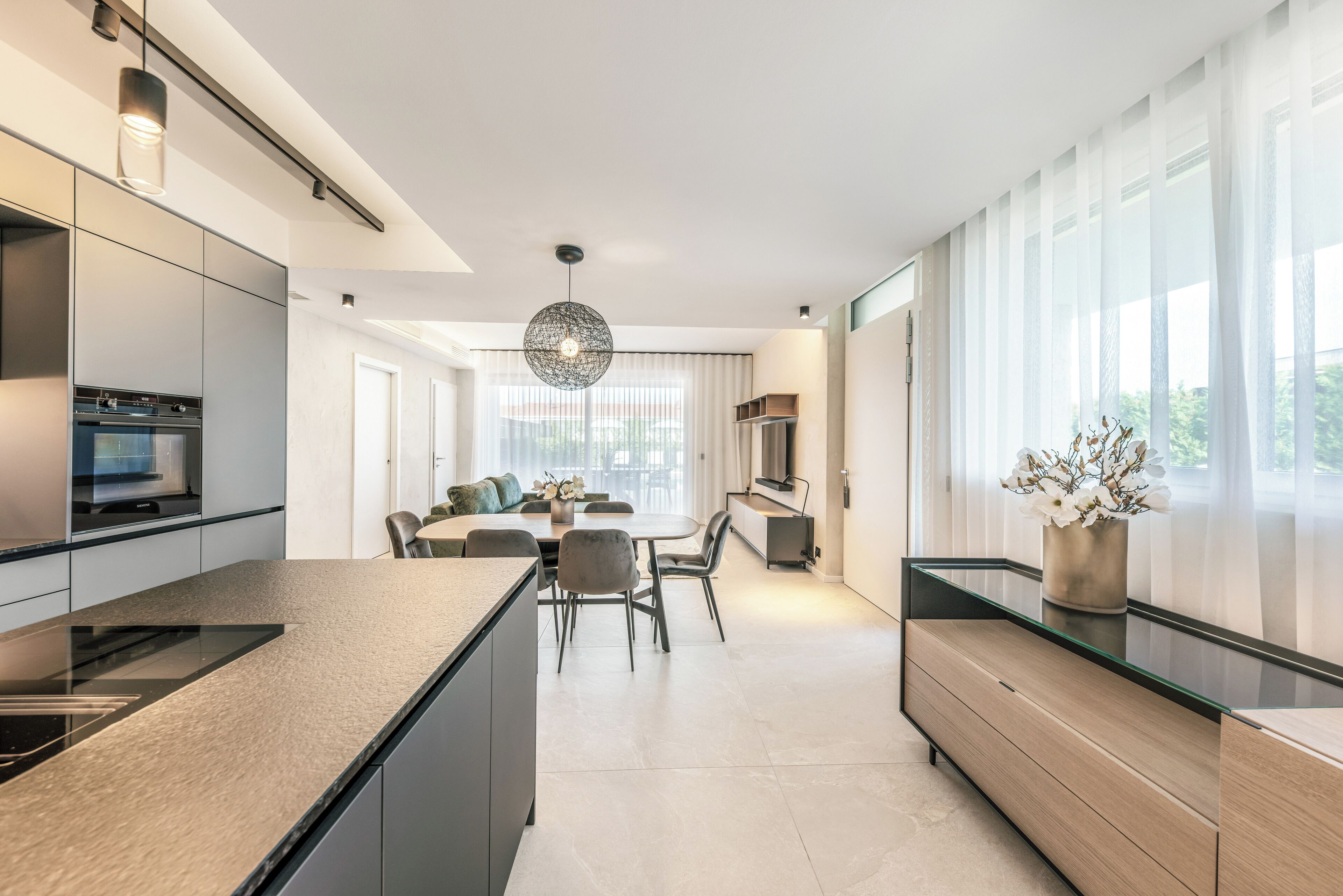 Uma cozinha moderna com ilha central, área de refeições com cadeiras e uma grande janela com cortinas leves.