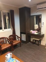 Quarto de hotel com uma mesa de jantar de madeira, duas cadeiras de madeira, um pequeno frigorífico, uma secretária com cadeira e um espelho grande.