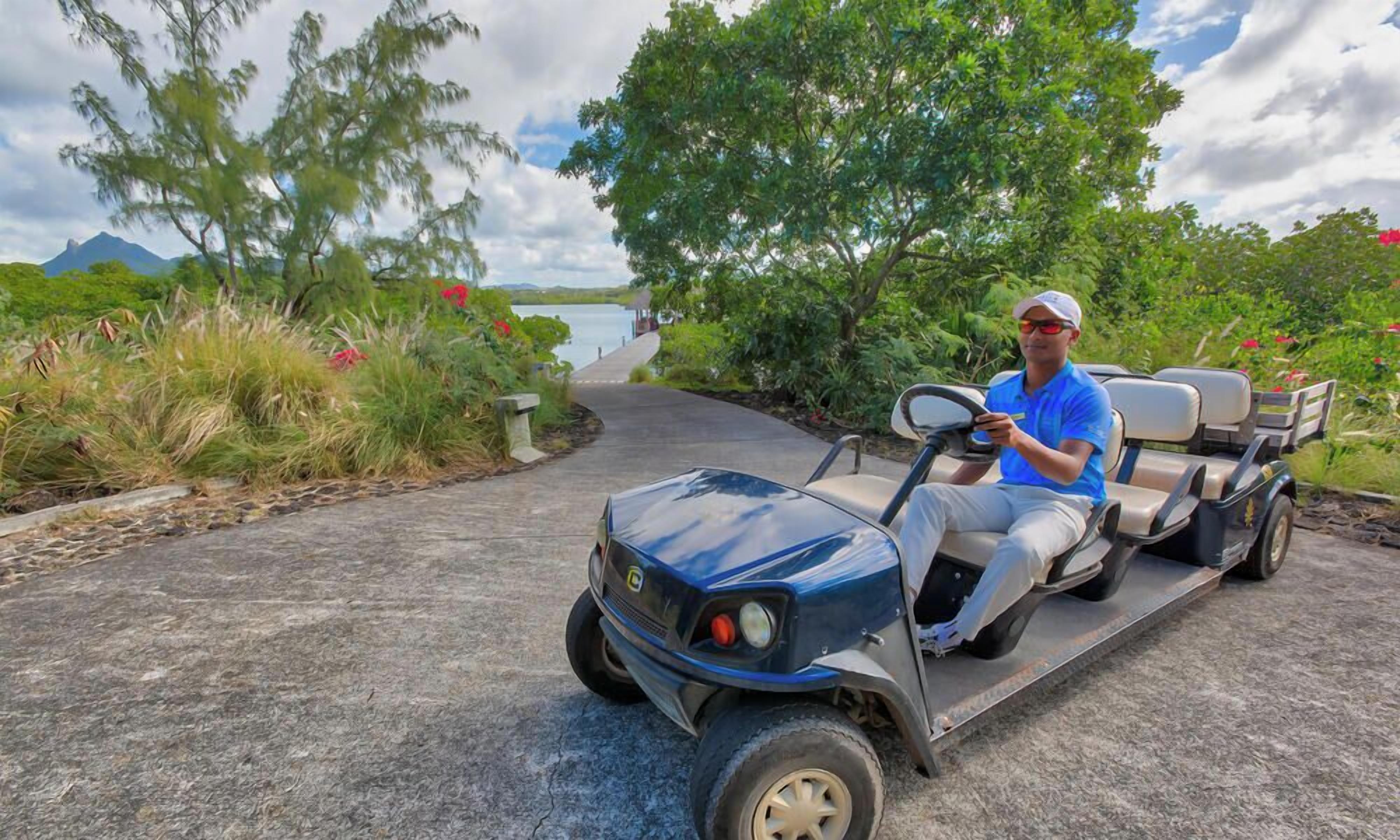 Um homem conduz um carrinho de golfe por um caminho pavimentado, rodeado por vegetação e um corpo d'água.