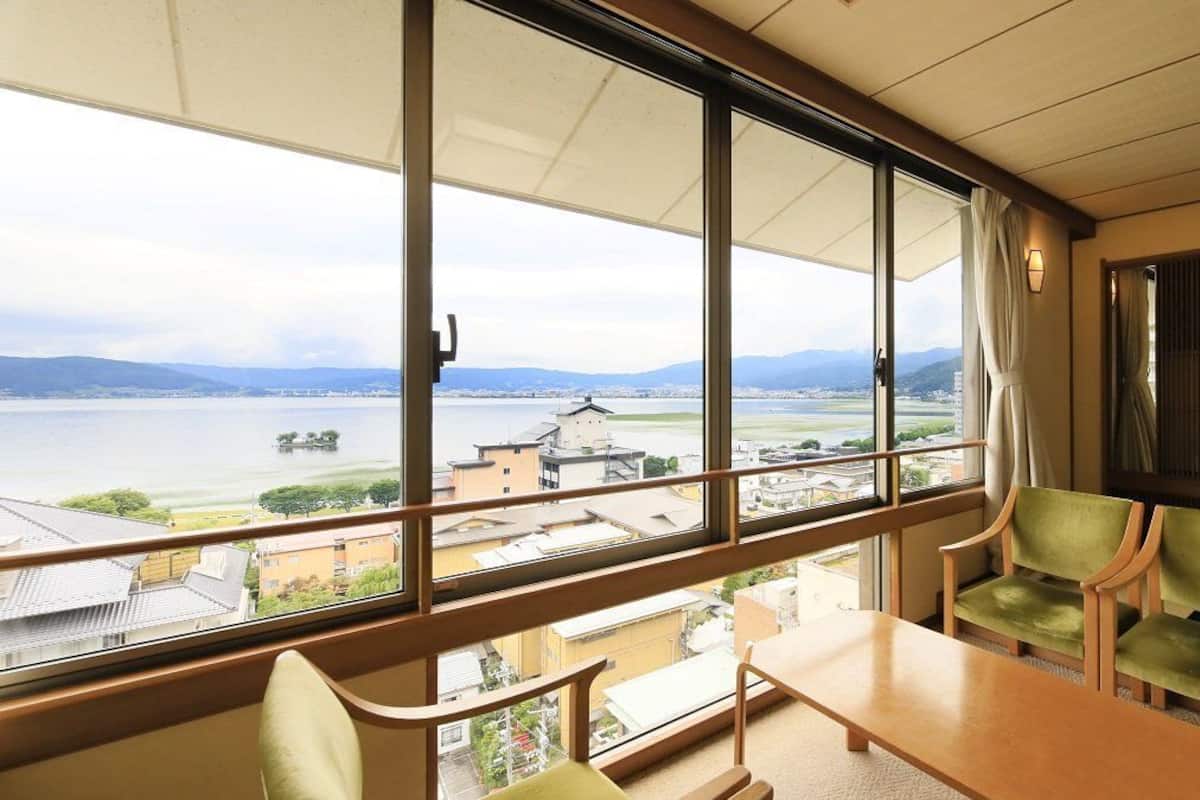 Quarto com uma grande janela, uma mesa de madeira, duas cadeiras verdes e vista para a água e edifícios.