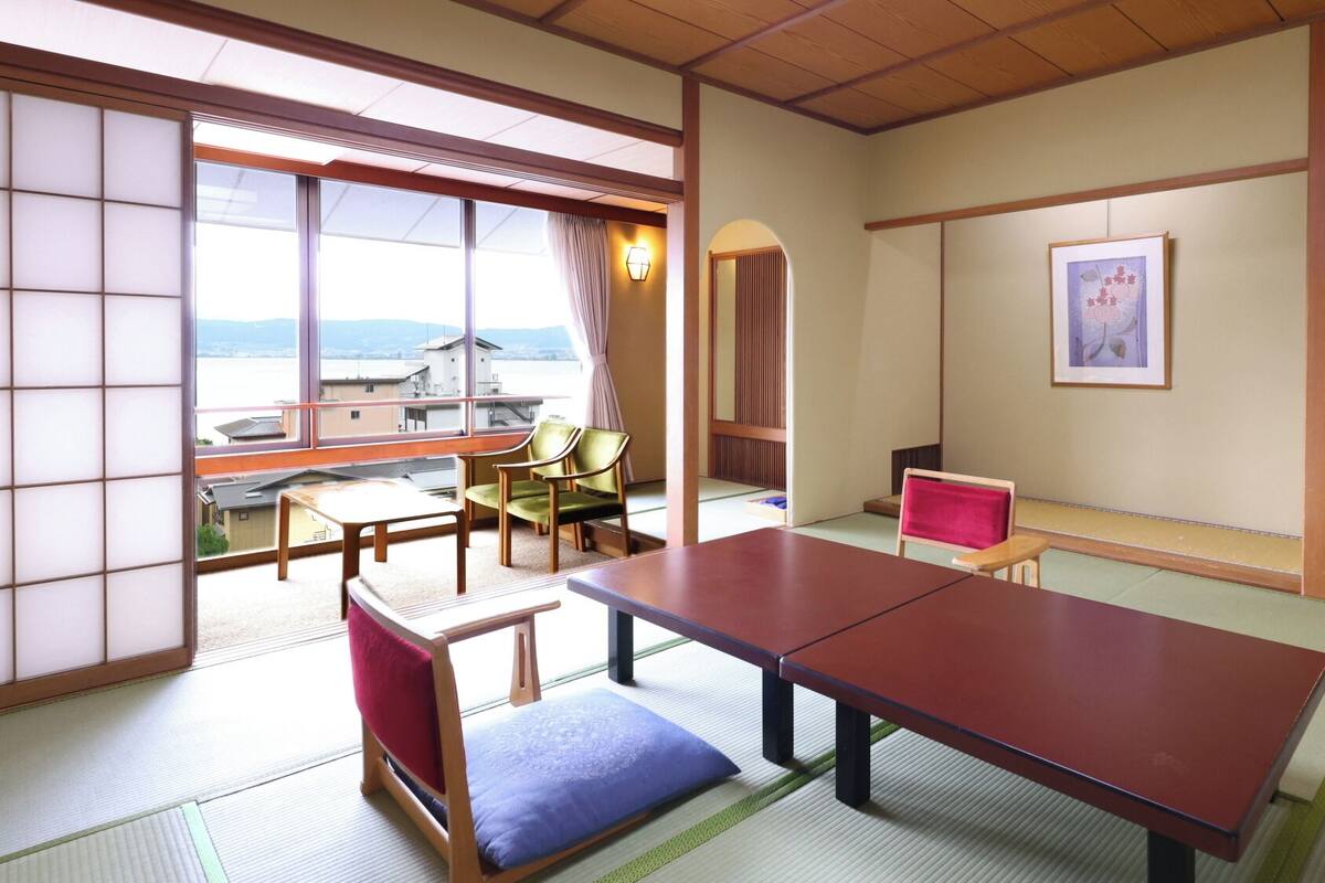 Quarto japonês tradicional com piso de tatame, uma mesa baixa, cadeiras e vista através de amplas janelas.