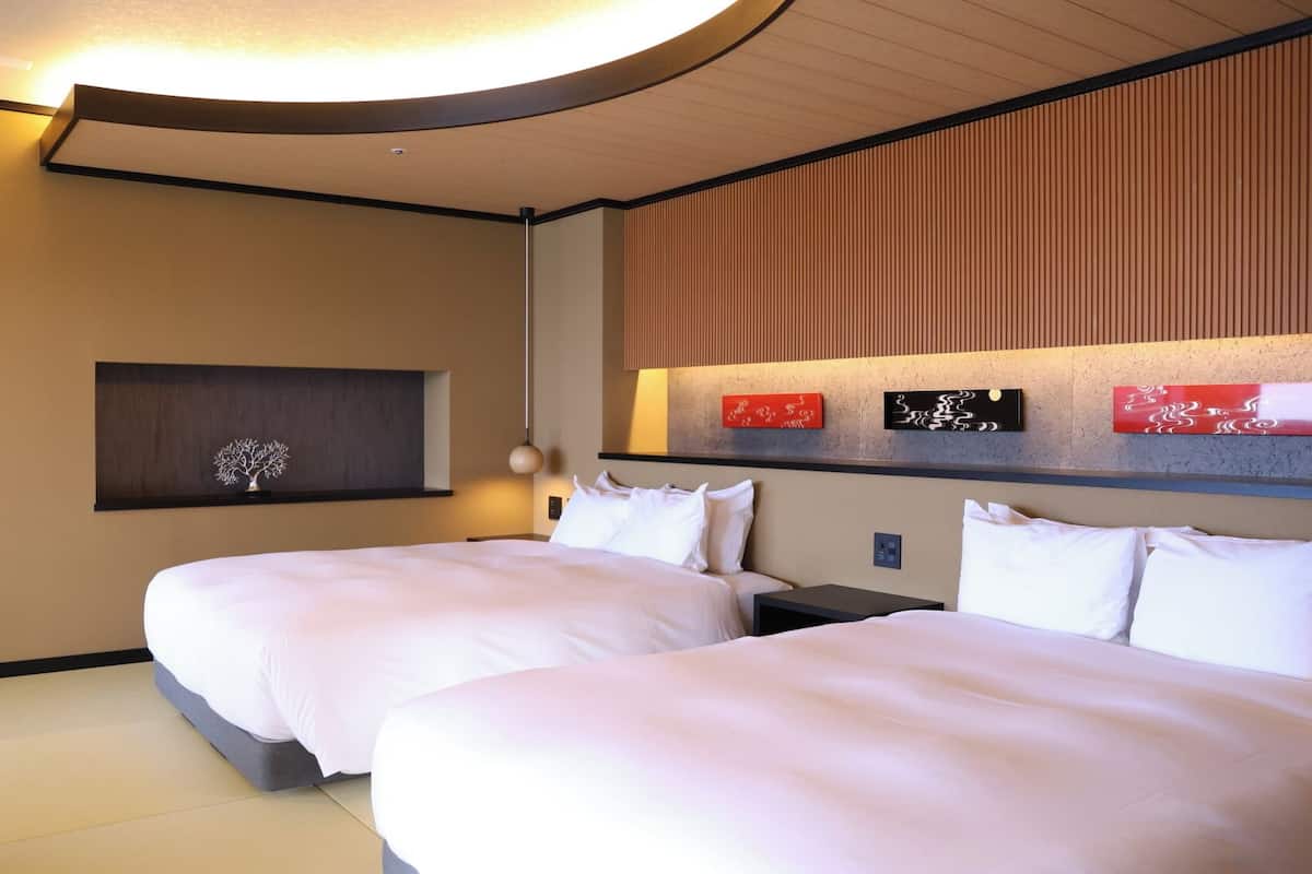 Quarto de hotel com duas camas, uma televisão presa na parede, um pequeno prateleira com um objeto decorativo e um candeeiro no teto.