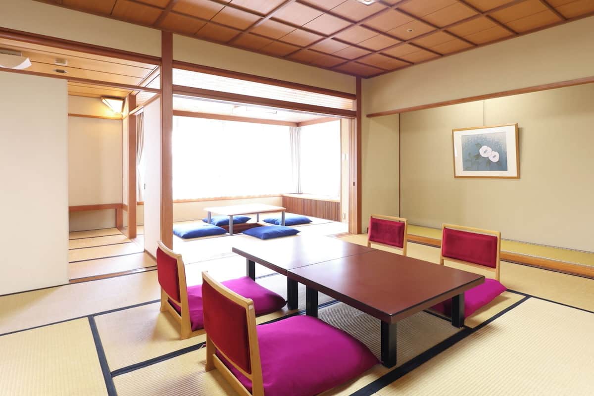 Um espaço com tatames, uma mesa baixa e cadeiras, uma janela grande e um quadro na parede.