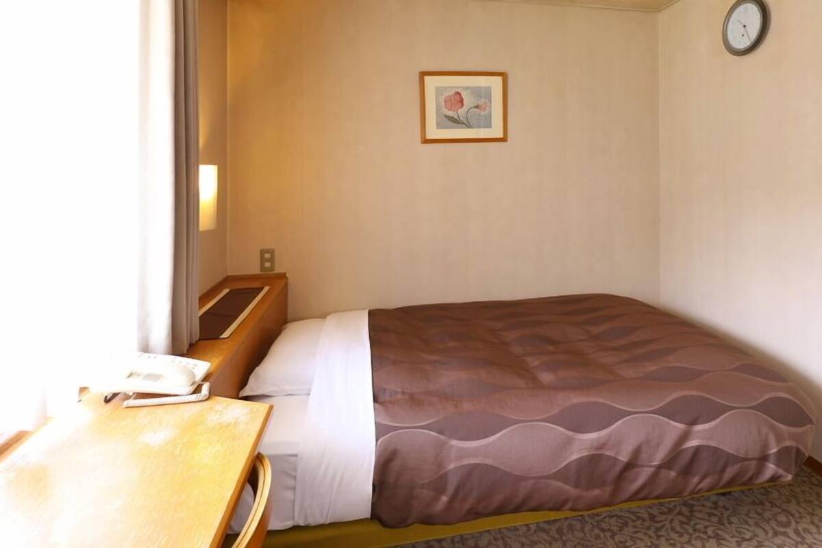 Quarto de hotel pequeno com uma cama de solteiro, uma escrivaninha de madeira, um relógio de parede e um quadro emoldurado.