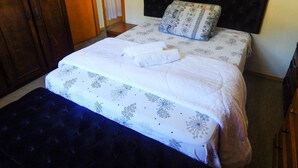 Uma cama bem arrumada com colcha estampada e travesseiros, um guarda-roupa de madeira e um banquinho escuro.