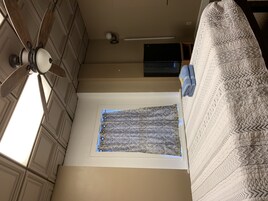 Quarto com ventoinha no teto, janela com cortinas estampadas, cama com colcha e um frigorífico.