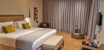 Quarto de hotel com uma cama grande, mesa de cabeceira com abajur, mesinha redonda com um vaso e cortina.