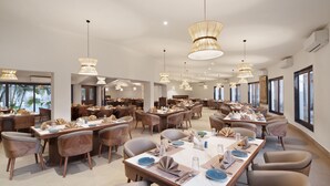 Uma sala de jantar ampla, com várias mesas e cadeiras, luminárias pendentes grandes e janelas amplas que oferecem luz natural.