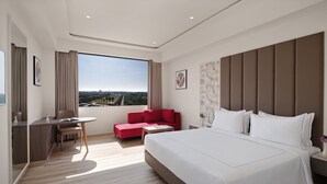 Quarto de hotel moderno com uma cama grande, uma escrivaninha, um sofá vermelho e vista para a cidade.