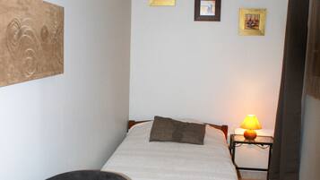 Quarto pequeno e estreito com uma cama de solteiro, uma cadeira e uma mesa de cabeceira com abajur. Há três quadros pendurados na parede.