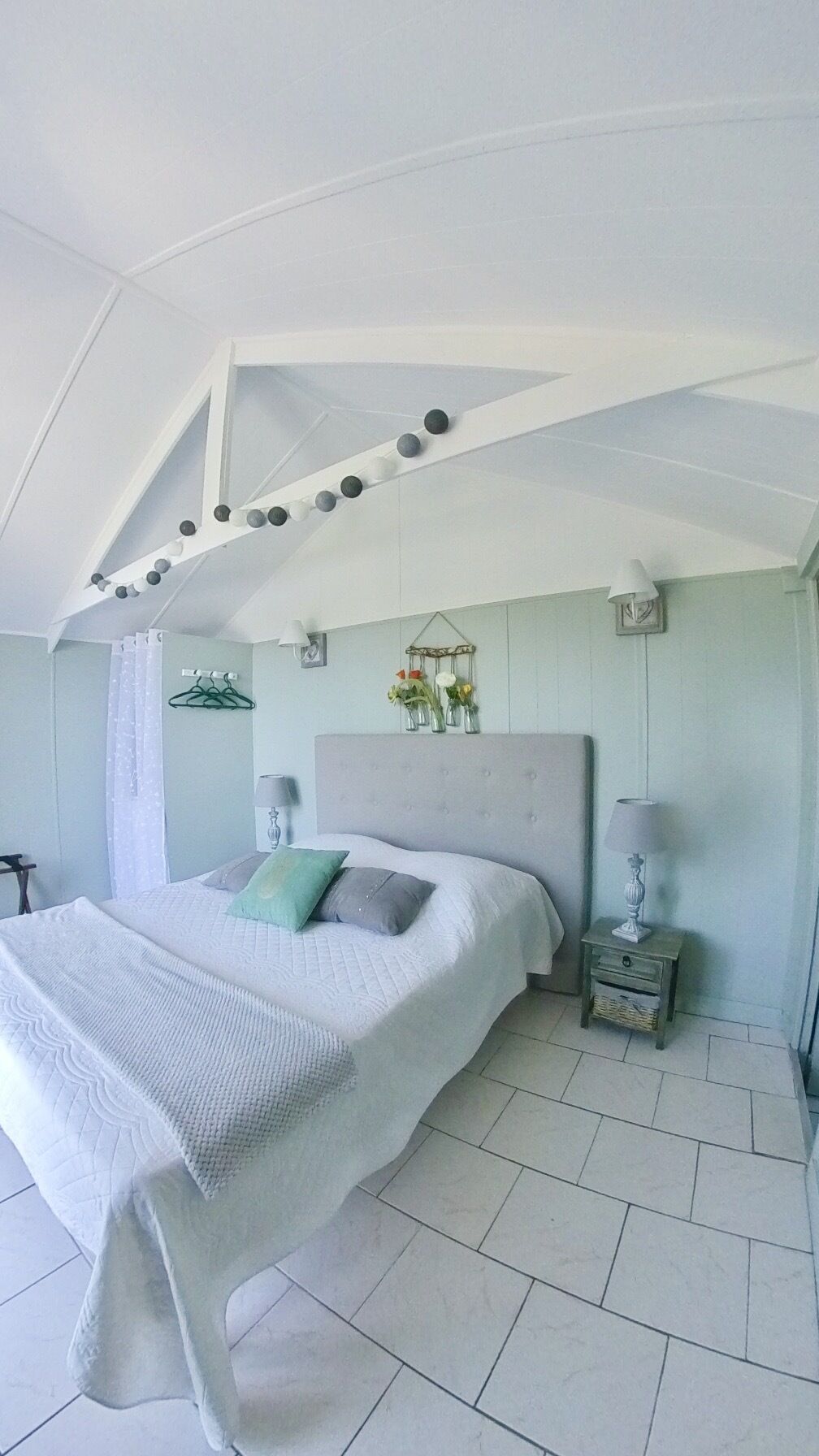 Quarto com cama grande, cabeceira, mesa de cabeceira com abajur e prateleira na parede com objetos decorativos.
