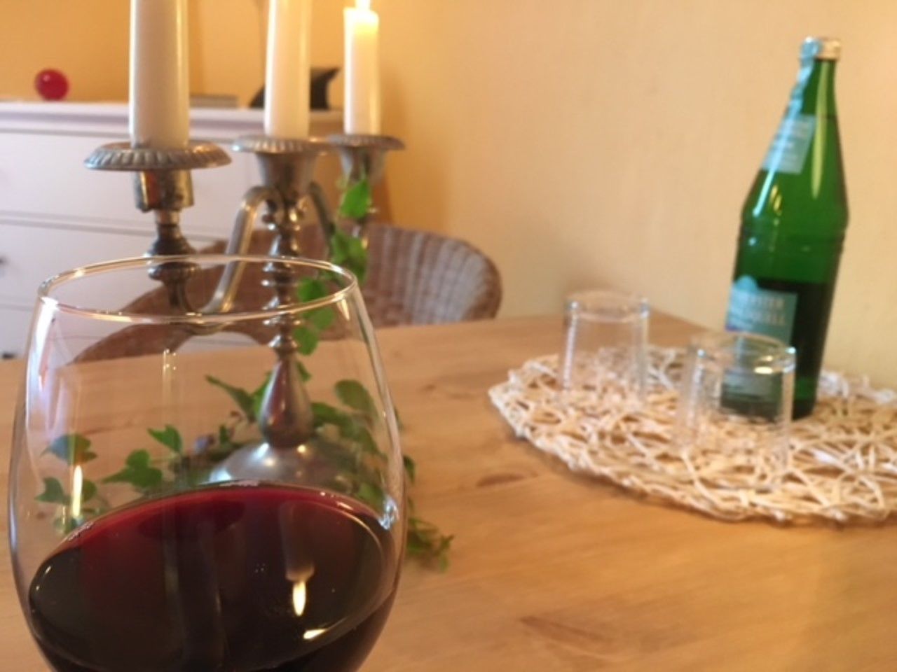 Um copo de vinho tinto sobre uma mesa de madeira com um jogo americano de renda, velas e uma garrafa de cerveja ao fundo.