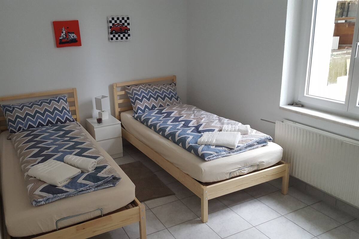 Quarto duplo com duas camas de solteiro, uma pequena mesa de cabeceira com luminária e uma janela com vista.
