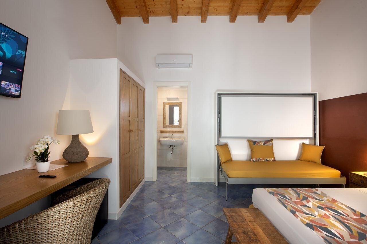 Quarto de hotel moderno com cama, escrivaninha, luminária, planta em vaso e vista para o banheiro.