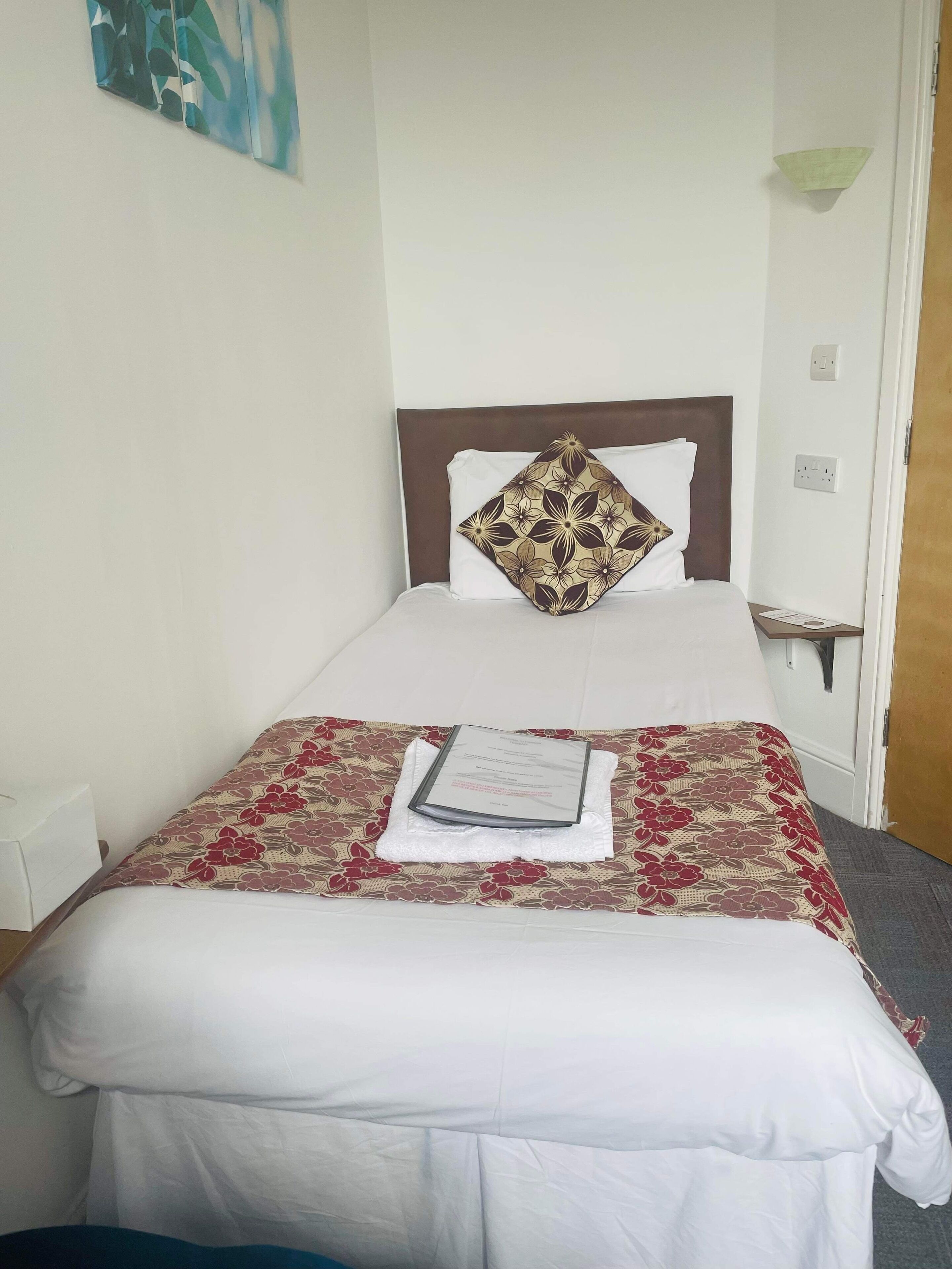 Uma cama bem arrumada com colcha estampada em floral, um travesseiro decorativo e um pequeno livreto sobre a cama.