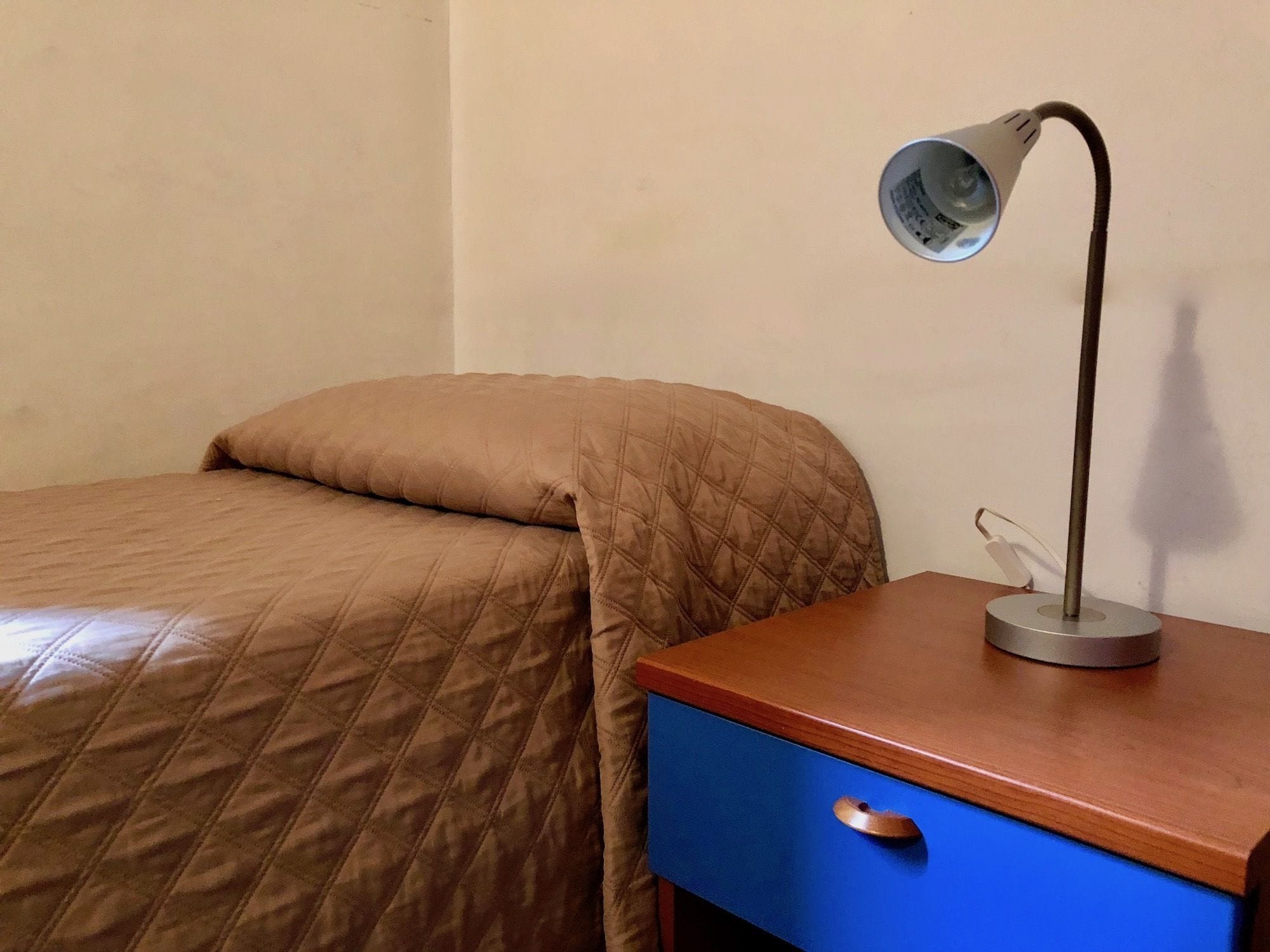 Uma cama de solteiro com um cobertor acolchoado castanho, uma mesa de cabeceira de madeira com uma gaveta azul e um abajur sobre a mesa de cabeceira.