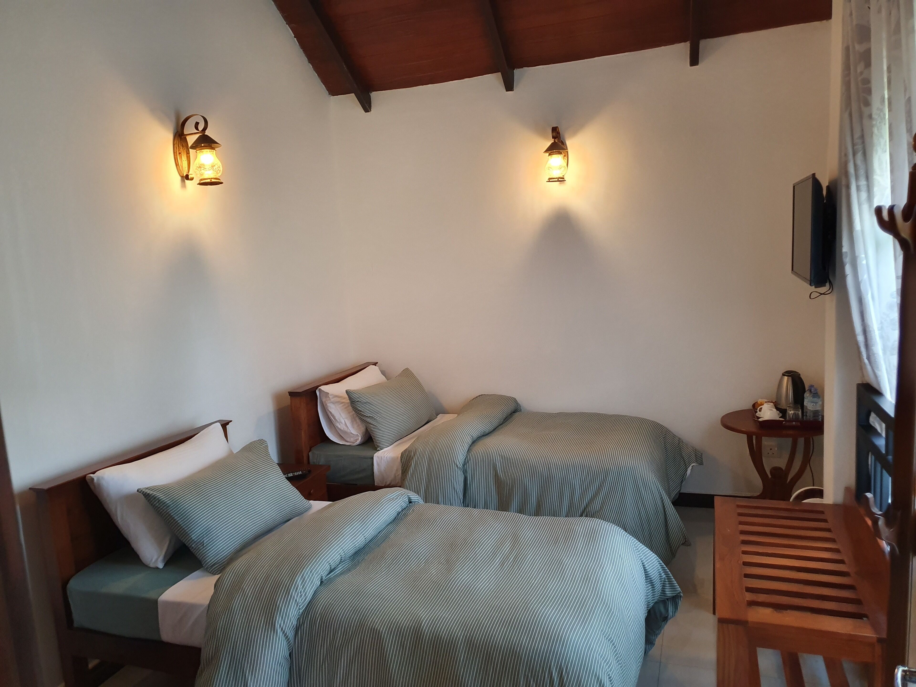 Quarto com duas camas, cabeceira de madeira, mesa de cabeceira com chaleira e duas luminárias de parede.