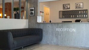 Uma área de recepção com um sofá cinzento, um balcão de recepção com a inscrição "RECEPTION", um expositor de chaves fixado na parede e um quadro emoldurado.
