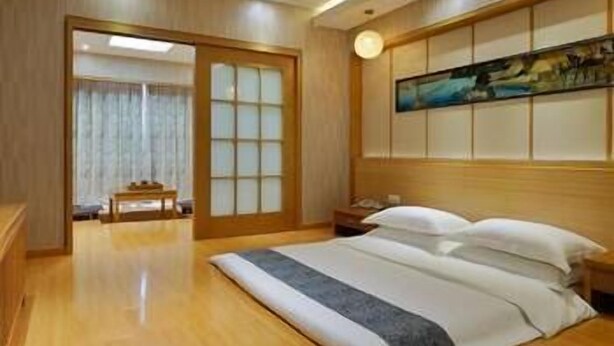 Quarto de hotel com piso de madeira, cama com roupa de cama branca, mesa de cabeceira, quadro na parede e porta deslizante para outro quarto.