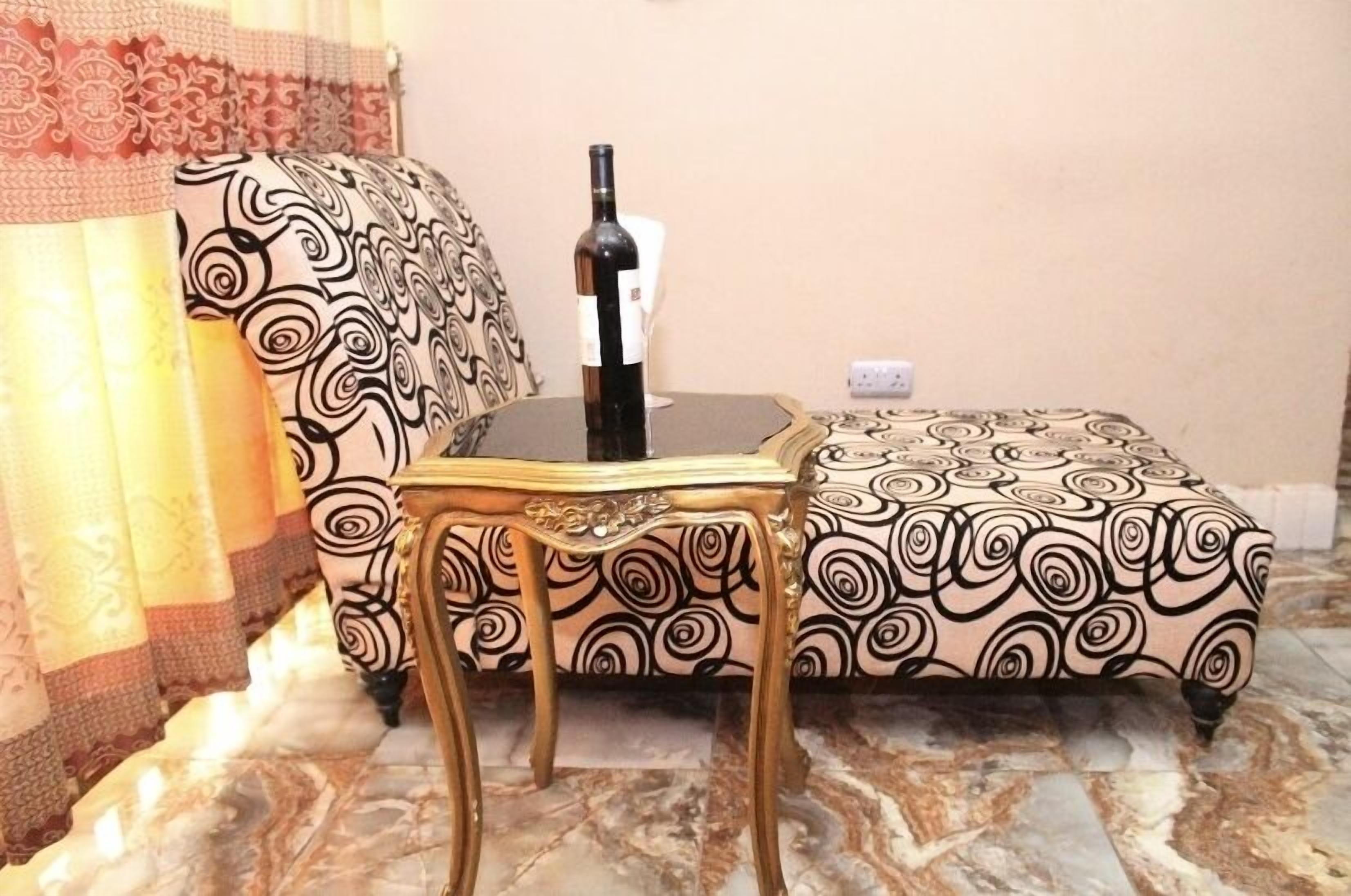 Um quarto com um sofá estampado, uma mesinha com uma garrafa de vinho e um banquinho estofado.