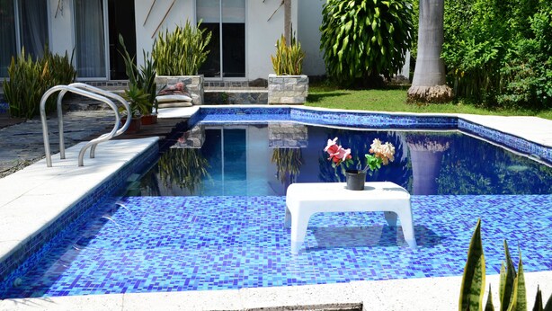 Uma piscina com design em mosaico azul, uma espreguiçadeira branca e plantas em vasos ao redor da área da piscina.