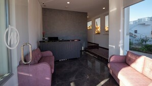 Um lobby moderno com um balcão de recepção, dois sofás, uma janela grande e uma escadaria para um andar superior.